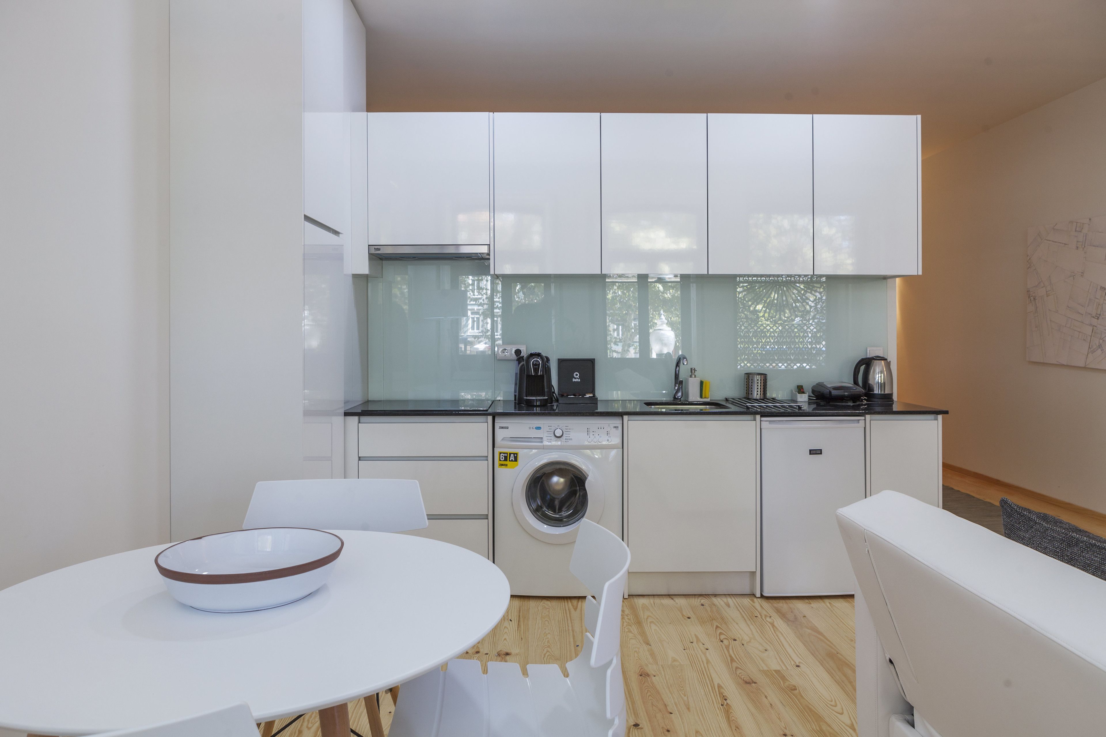 Uma cozinha compacta com armários brancos, uma máquina de lavar e uma pequena área de refeições com uma tigela sobre a mesa.
