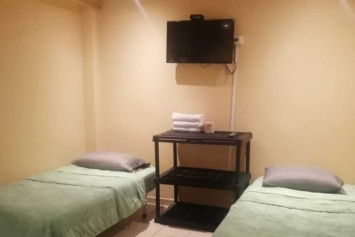 Quarto com duas camas de solteiro, cada uma com travesseiro e manta verde. Há uma mesinha com uma televisão em cima e uma pilha de toalhas dobradas.