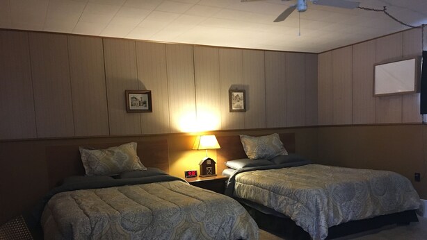 Quarto de hotel com duas camas, uma mesa de cabeceira com abajur, um relógio e uma pequena mesa com um jornal.
