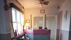 Quarto de hotel com balcão de recepção, uma cadeira, luminária de mesa, ventilador de teto e janela com persianas.