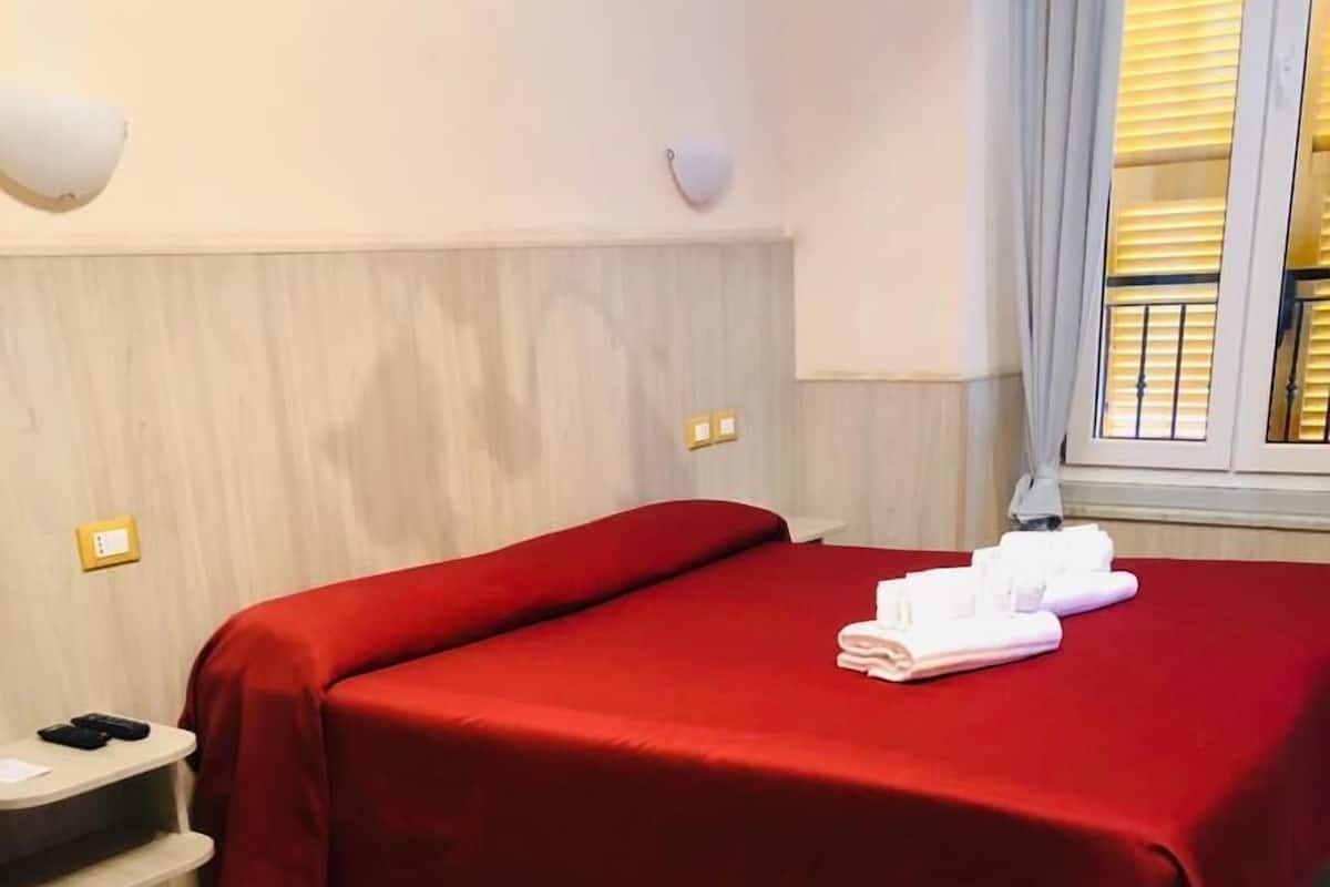 Quarto de hotel com cama, mesa de cabeceira, luminária e janela com persianas.