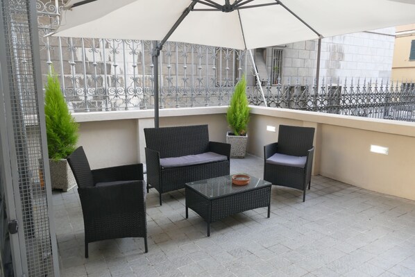 Varanda com móveis de vime, mesa de vidro e plantas em vasos.