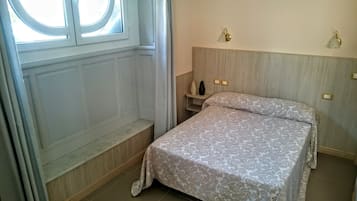 Quarto pequeno e bem arrumado, com cama, mesa de cabeceira, janela e luminária fixada na parede.