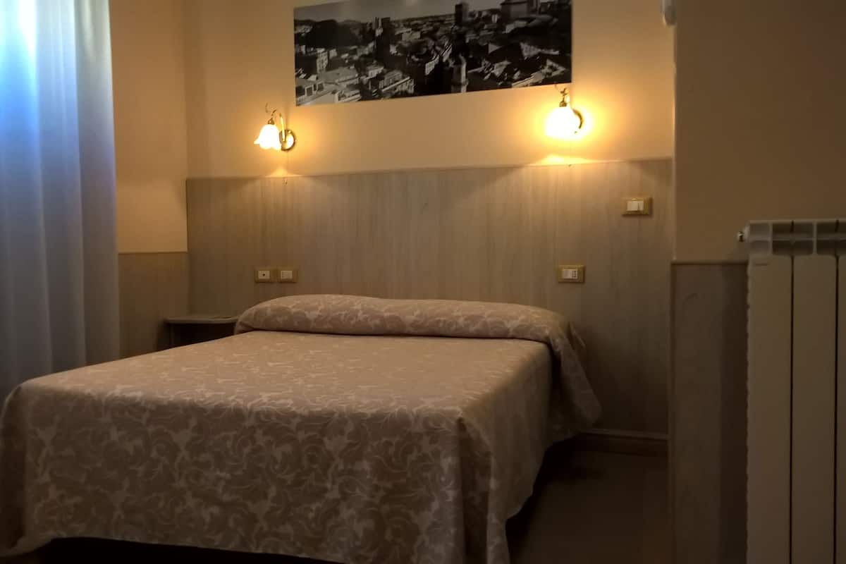 Quarto de hotel com uma cama, duas luminárias fixadas na parede, um radiador e uma pintura com vista para a cidade acima da cama.