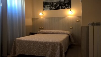 Quarto de hotel com uma cama, duas luminárias fixadas na parede, um radiador e uma pintura com vista para a cidade acima da cama.