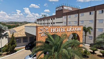 Entrada de hotel com uma placa indicando 'HOTEL LIGUORI', rodeada por palmeiras e carros estacionados.