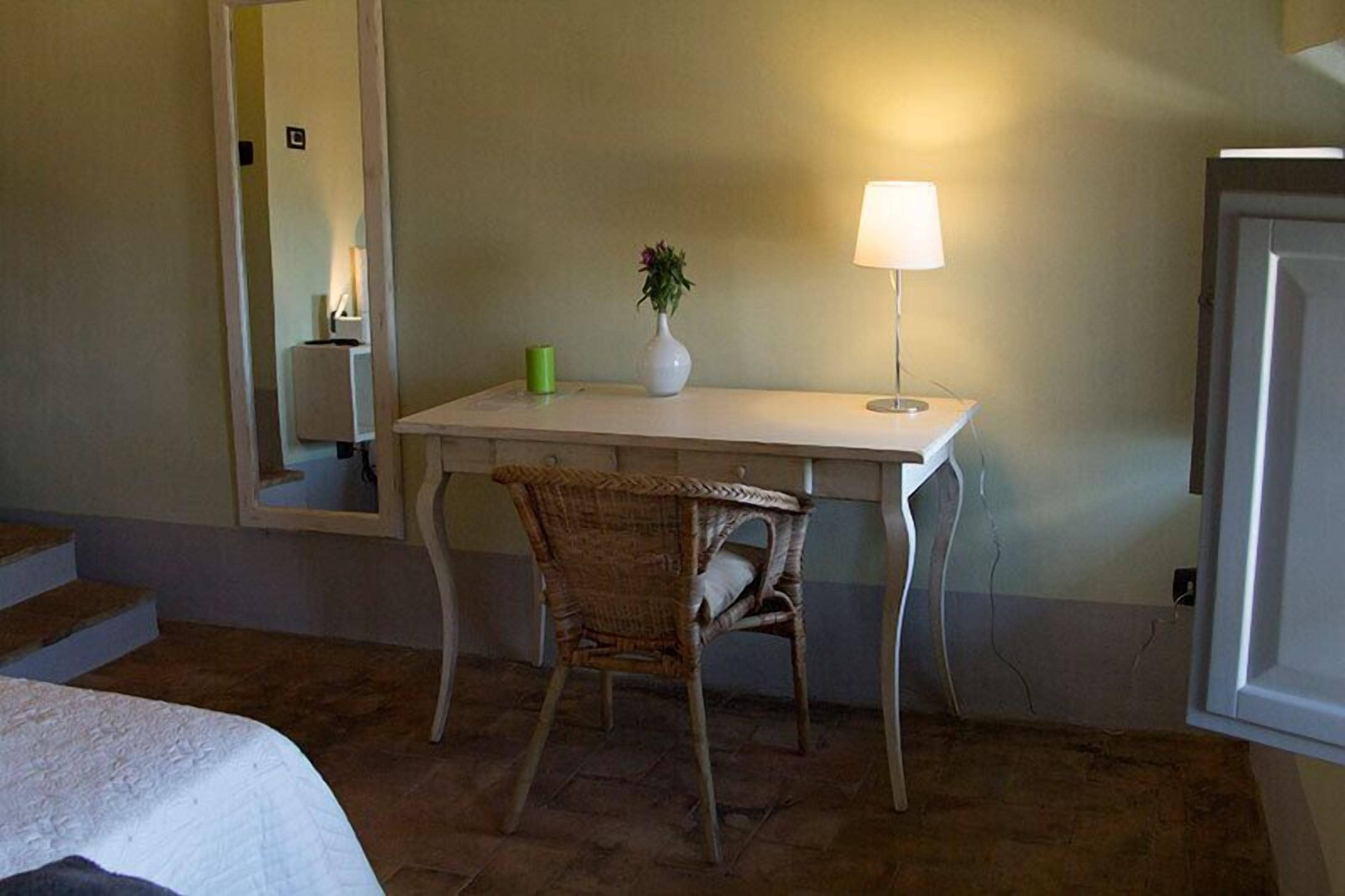 Quarto com cama, escrivaninha com luminária, cadeira, vaso com flores e espelho.