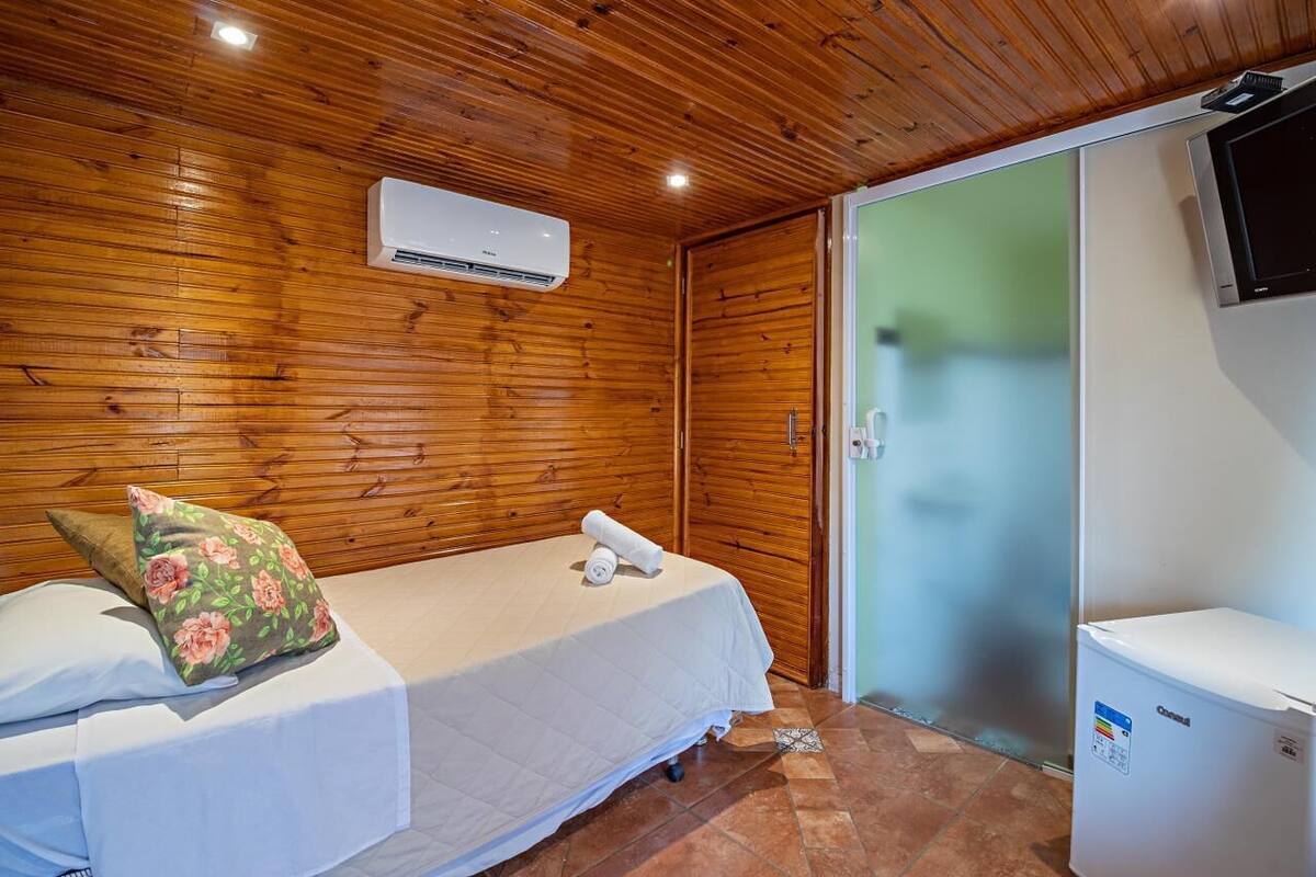 Quarto com paredes de madeira, cama com colcha branca e almofada floral, ar condicionado branco, televisão e geladeira.