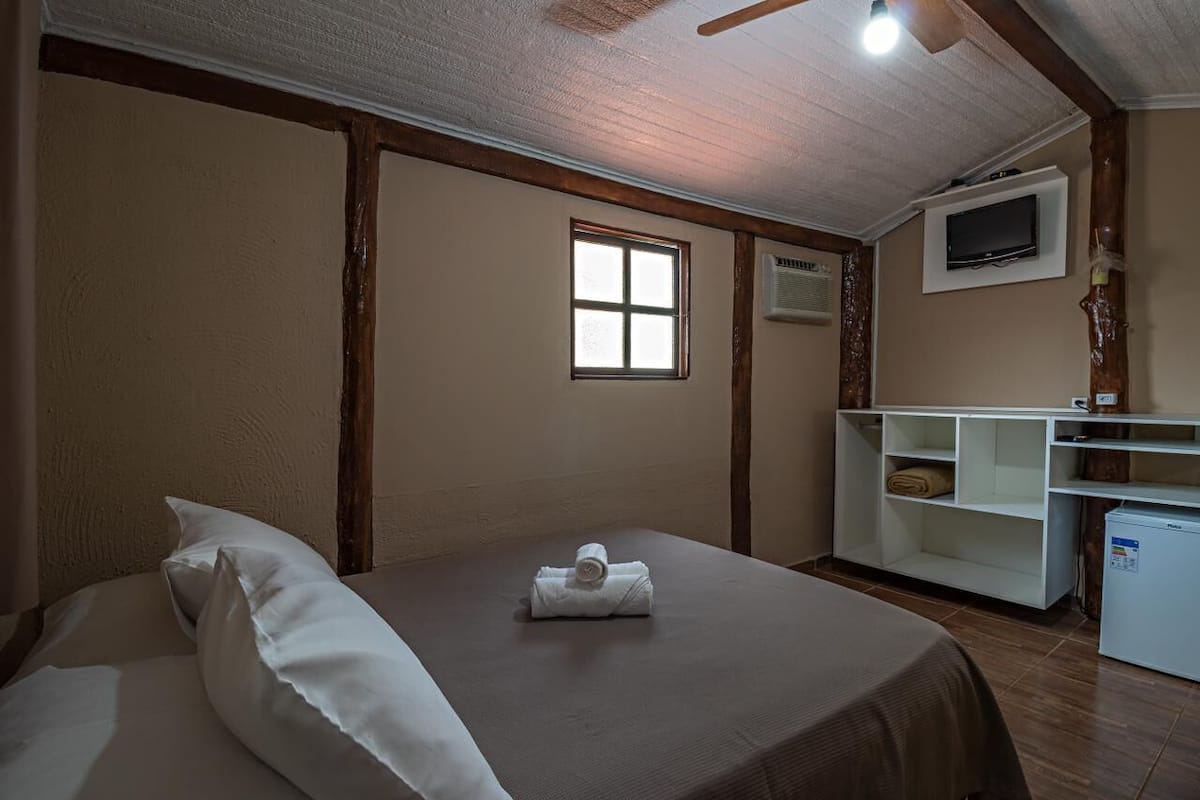 Quarto de hotel com uma cama, uma janela pequena, uma televisão e um armário branco.