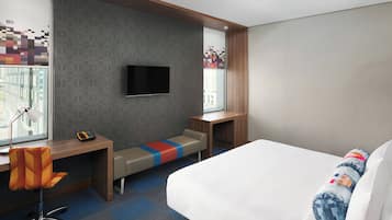 Quarto de hotel com cama, escrivaninha com cadeira, televisão na parede e janela com vista para edifícios.