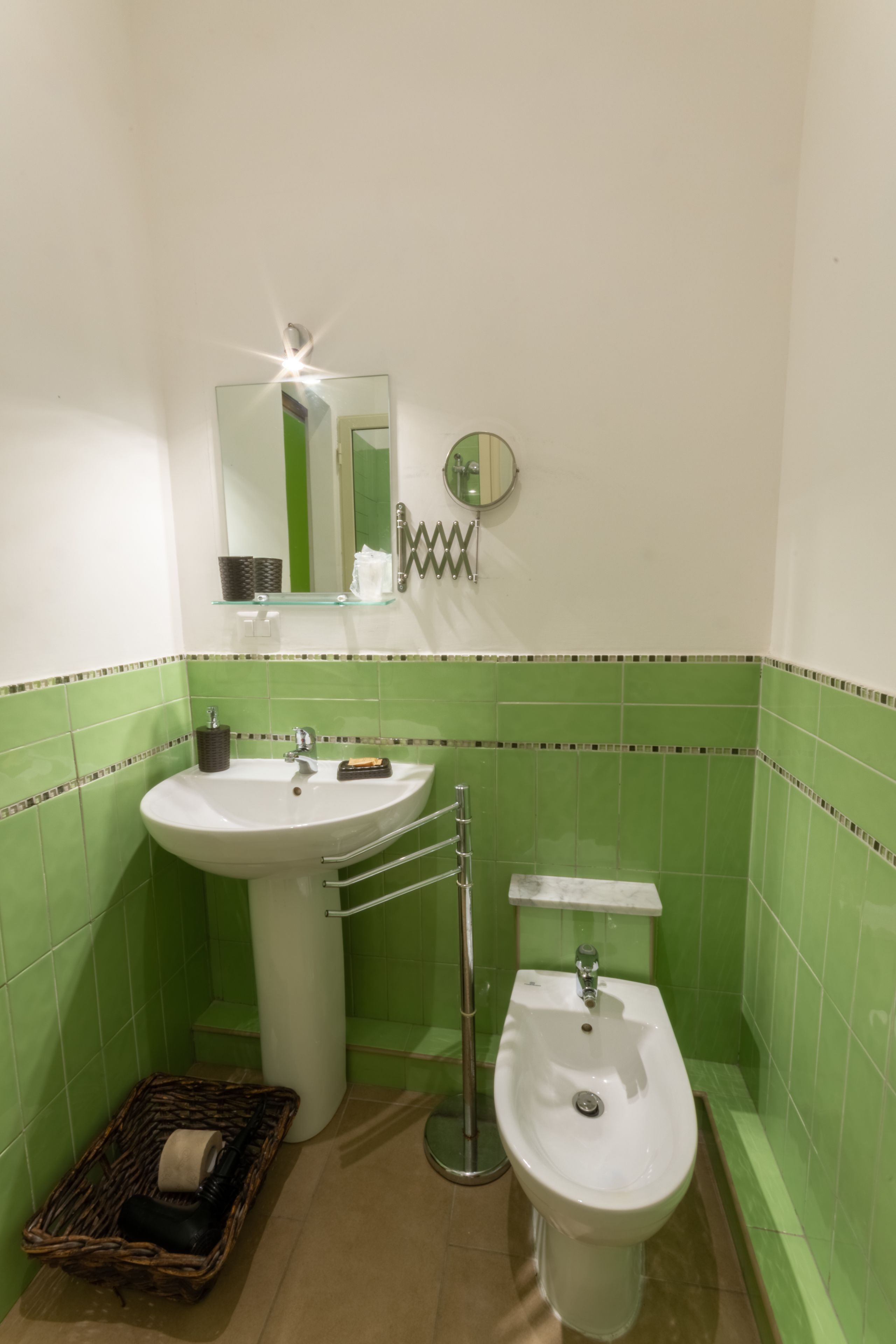 Casa de banho com lavatório branco, bidé, espelho e pequena prateleira com produtos de higiene pessoal.