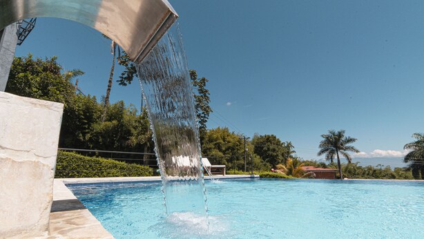 Uma piscina azul límpida com um elemento aquático, cercada por vegetação e um céu claro.