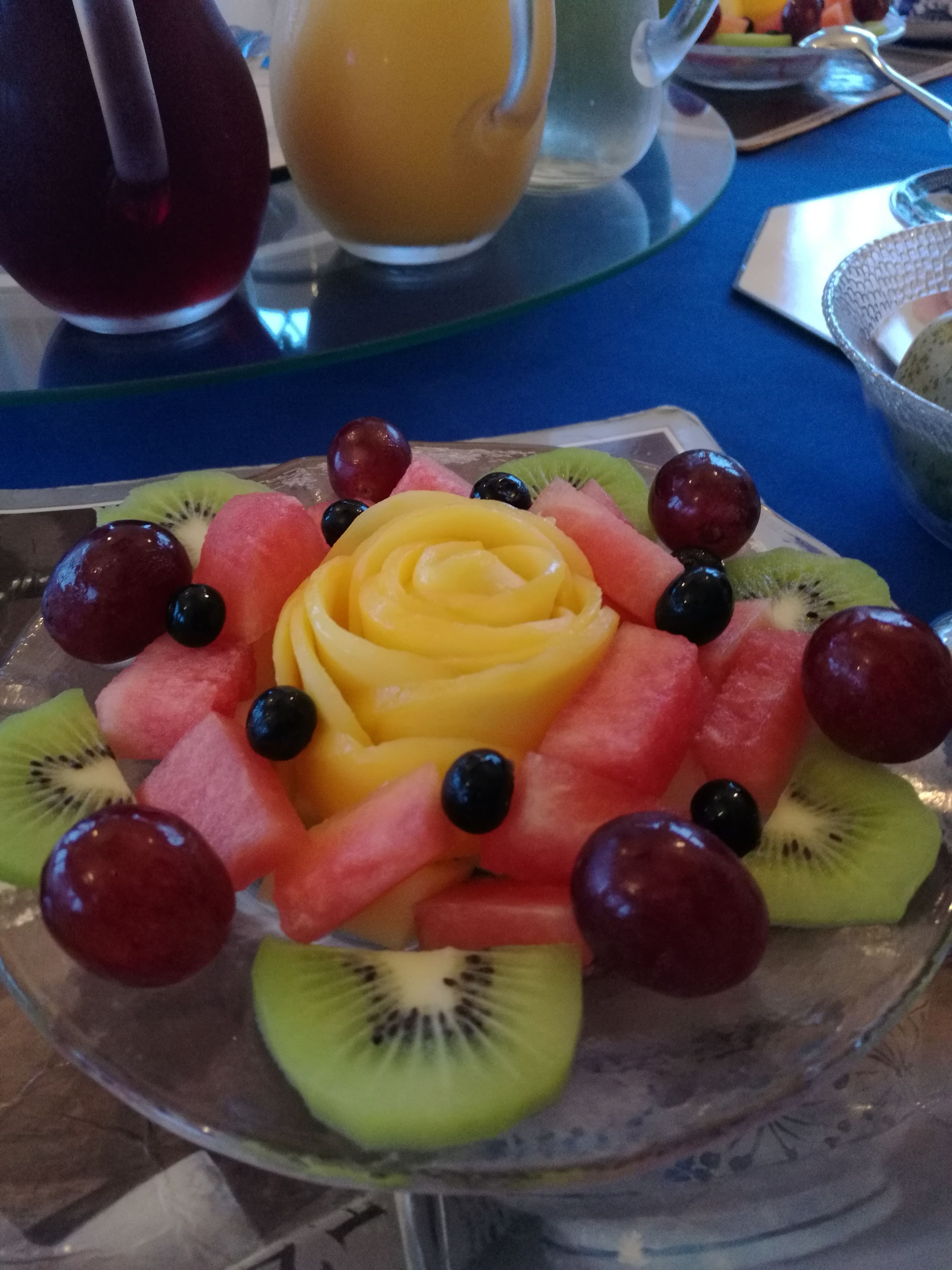 Uma travessa de frutas com melancia, kiwi, uvas e um mango no centro, sobre um prato de vidro.