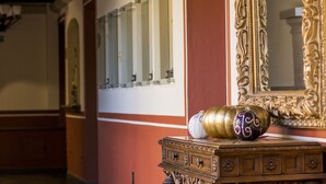 Um corredor com teto de madeira, piso de mármore, um espelho grande e ornamentado, e uma pequena mesa com objetos decorativos.