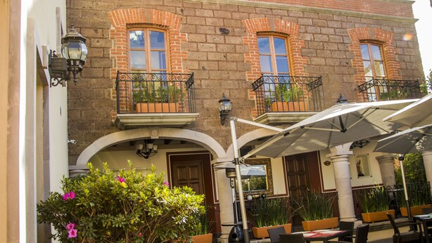 Um edifício de tijolos com três janelas, cada uma com uma varanda e vasos de plantas. Há mesas e cadeiras ao ar livre, e um guarda-sol branco oferece sombra.