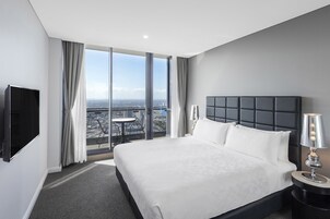 Quarto de hotel moderno com uma cama grande, televisão de tela plana, abajur e vista para o horizonte urbano através de janelas do chão ao teto.