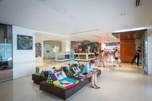 Um lobby moderno com área de estar, incluindo um sofá marrom com almofadas coloridas, balcão de recepção e parede de vidro.
