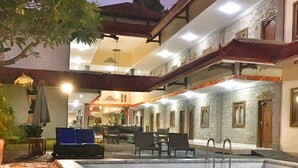 Piscina com deck de madeira e espreguiçadeiras, cercada por um edifício com várias varandas e telhado telhado.