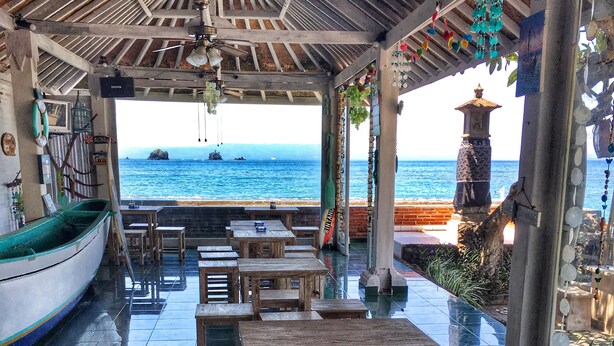 Uma área de estar coberta ao ar livre, com mesas e bancos de madeira, com vista para o mar.