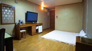 Quarto de hotel com piso de madeira, cama com roupa de cama branca, escrivaninha com televisão e uma pequena área de estar.