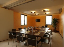 Uma sala de conferências com uma mesa comprida, cadeiras, uma televisão e um vaso de planta.