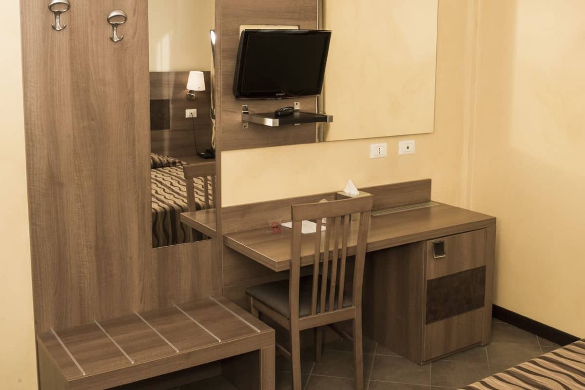 Quarto de hotel pequeno com escrivaninha de madeira, uma cadeira, uma cama com colcha estampada, um espelho e uma televisão.