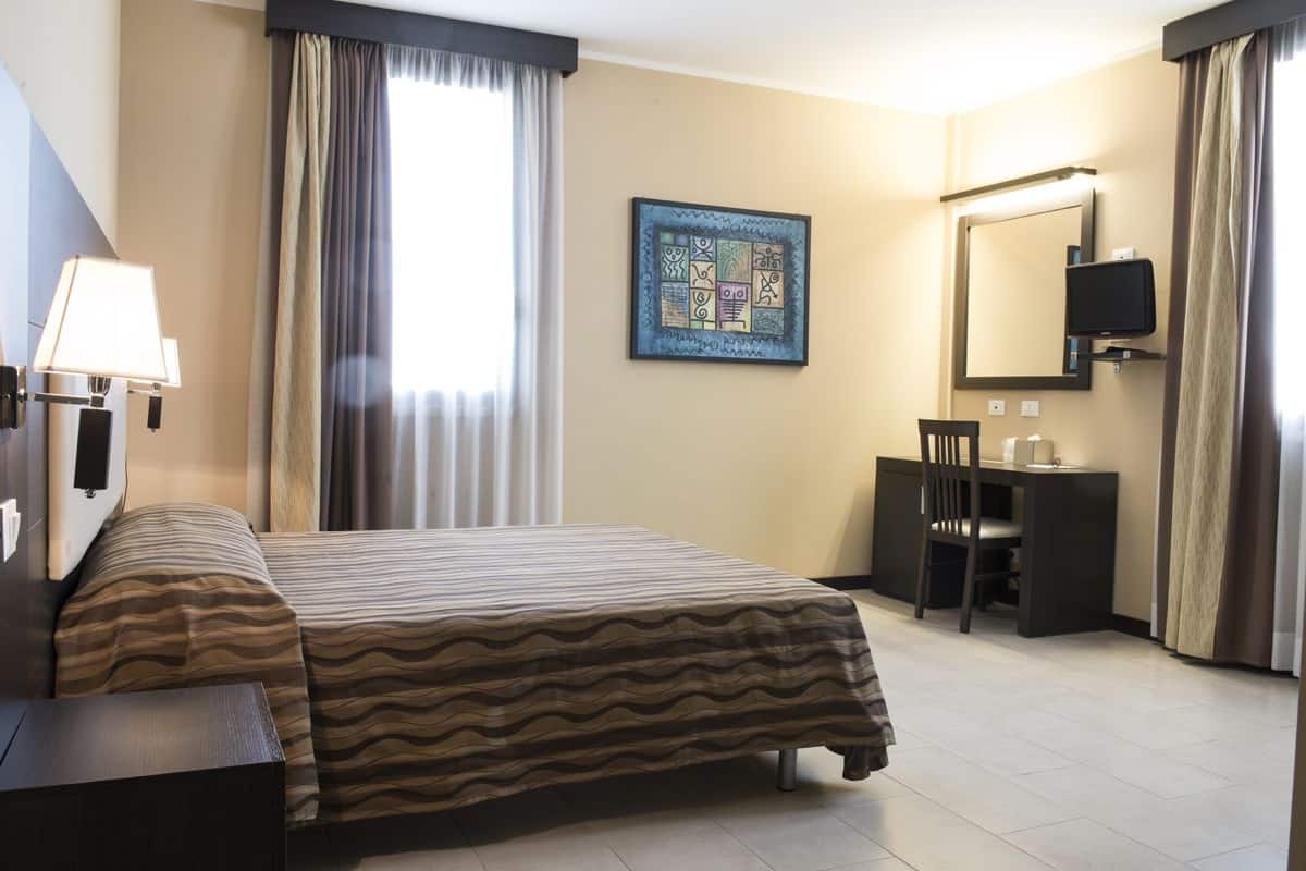 Quarto de hotel com cama, escrivaninha, cadeira, televisão e um quadro na parede.