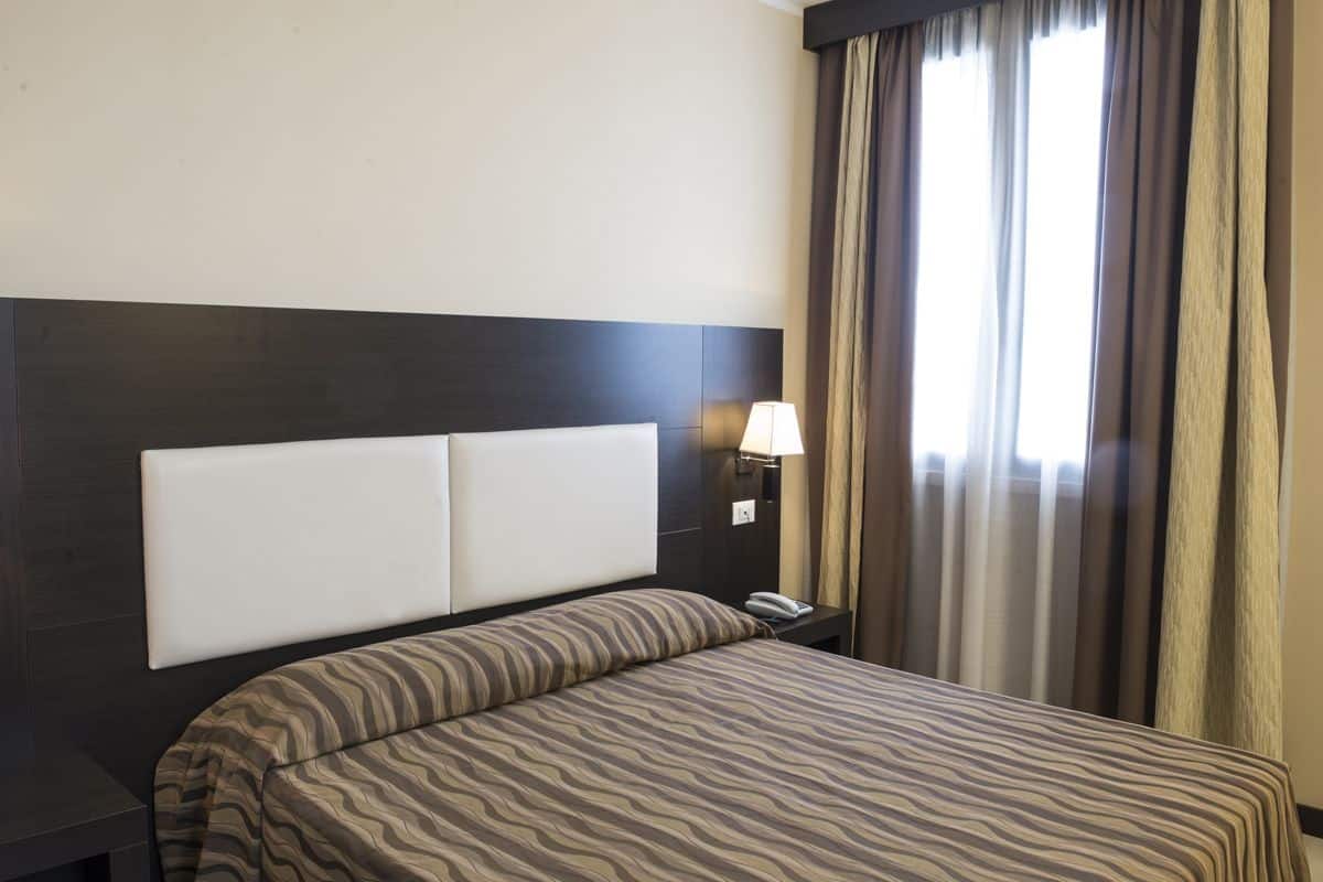 Quarto de hotel com uma cama, mesa de cabeceira, luminária e janela com cortinas.