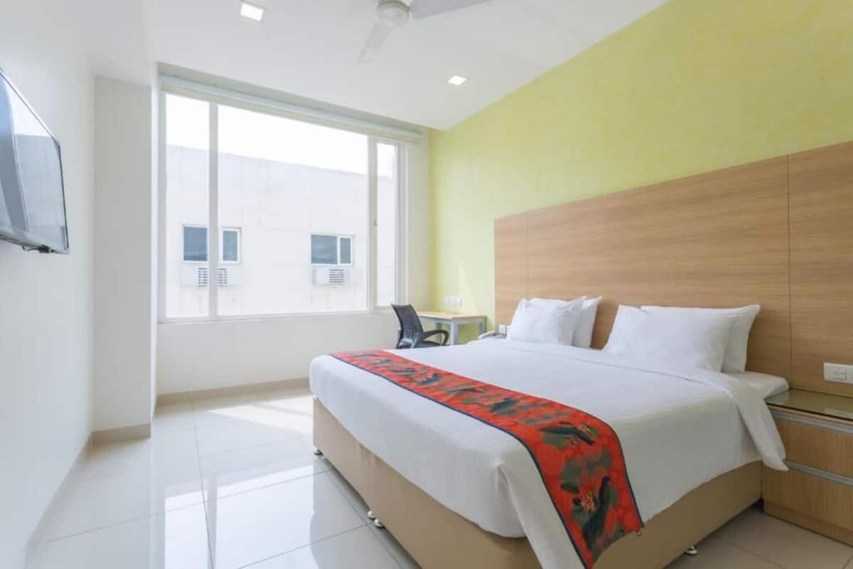 Quarto de hotel com cama, escrivaninha, cadeira, janela e televisão.