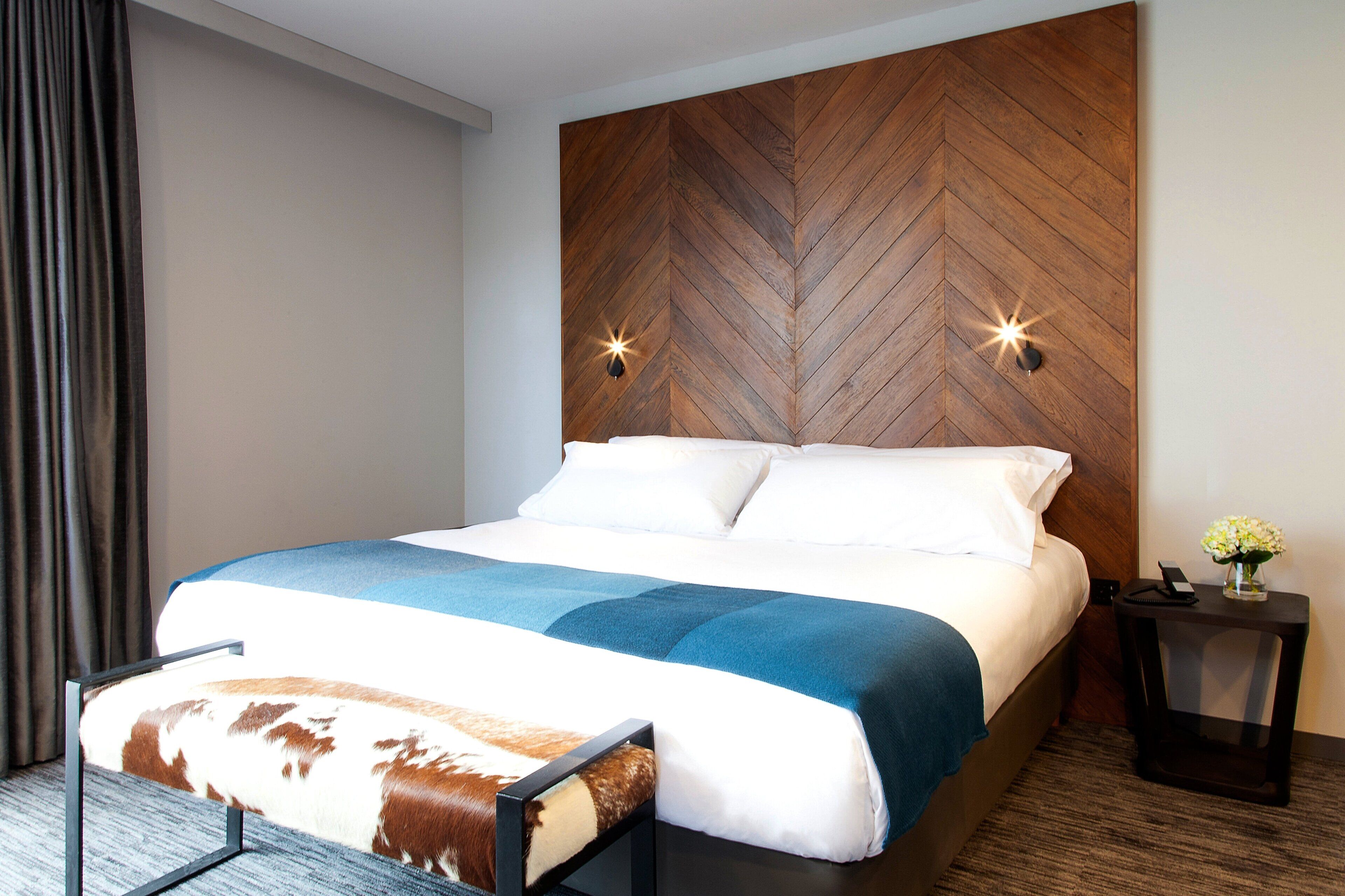 Quarto de hotel com uma cama grande, cabeceira de madeira, duas luzes fixadas na parede, mesa de cabeceira pequena com um telefone e um vaso de flores, e um cobertor marrom e branco na cama.