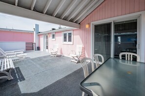 Um alpendre coberto com mesa e cadeiras, uma porta de correr e um edifício rosa ao fundo.
