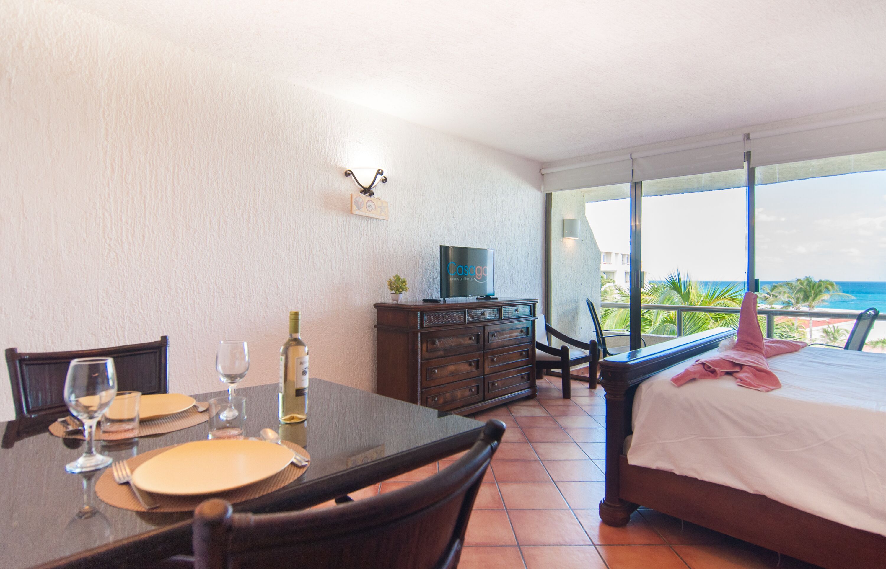 Quarto de hotel com cama, mesa de jantar posta para dois, televisão e vista para o oceano.