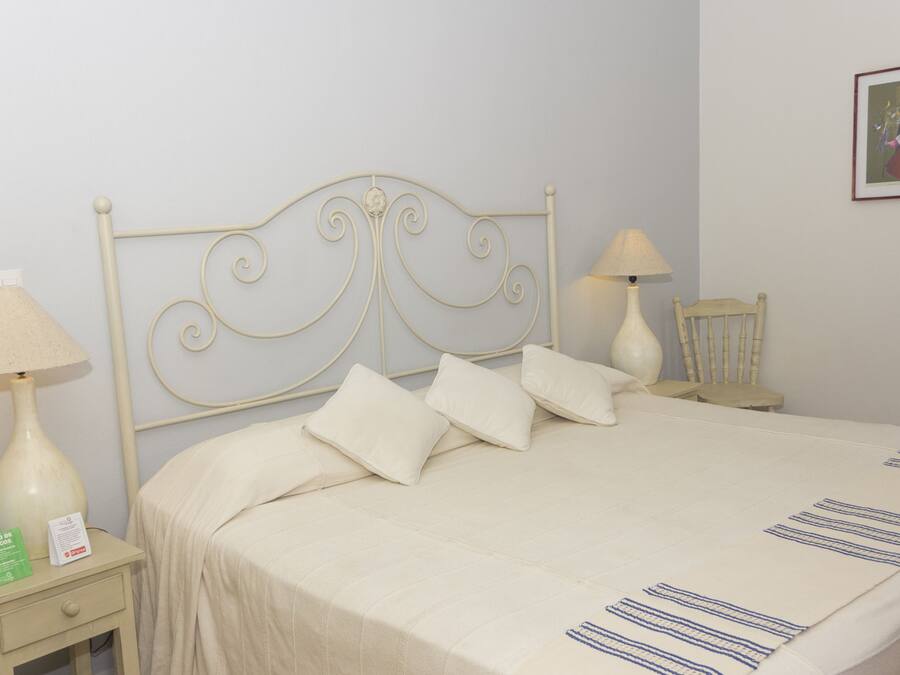 Cama bem arrumada com cabeceira, duas luminárias de cabeceira, uma cadeira e uma mesinha com abajur e placa.