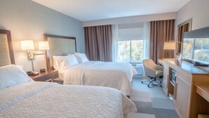 Quarto de hotel com duas camas, uma mesa de trabalho, televisão e janela com cortinas.