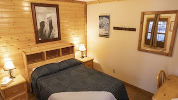 Quarto com painéis de madeira, uma cama, duas mesas de cabeceira com luminárias, um quadro na parede, uma janela com cortinas e uma pequena cadeira de madeira.