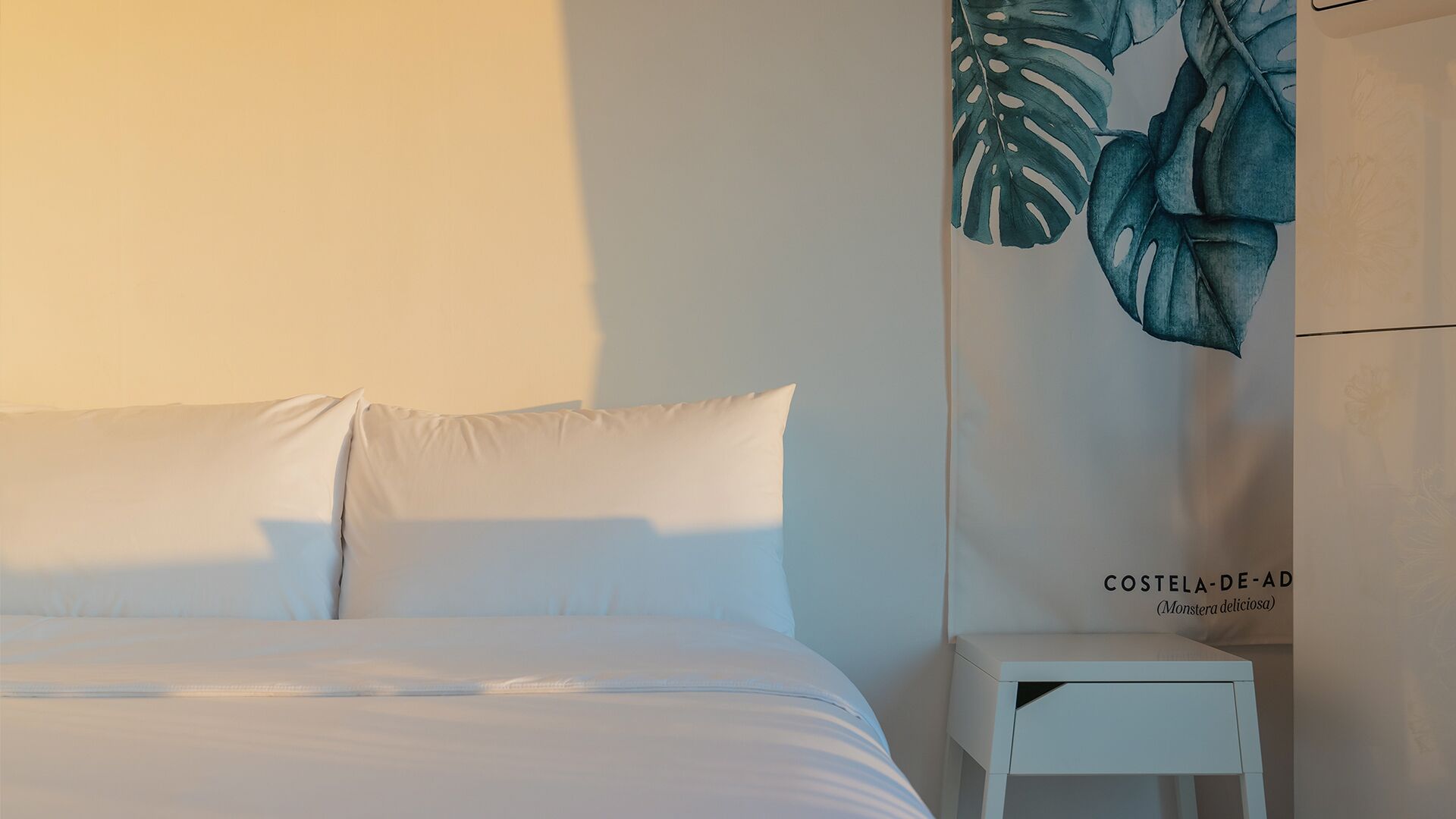 Uma cama bem arrumada com lençóis brancos, uma pequena mesa de cabeceira e uma parede com uma obra de arte de folha tropical.