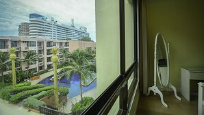 Quarto de hotel com vista para uma piscina, palmeiras e um edifício alto.