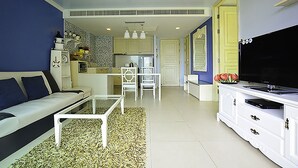 Uma sala de estar com sofá, mesa de centro e televisão sobre um móvel. Dá para ver uma cozinha ao fundo, com armários e geladeira.