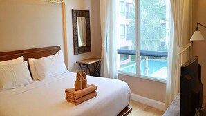Quarto de hotel com uma cama grande, uma mesinha, uma televisão e uma janela com vista para prédios e árvores.