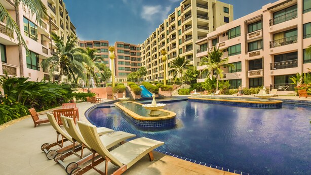 Área de piscina de hotel com espreguiçadeiras, uma fonte de água e palmeiras.