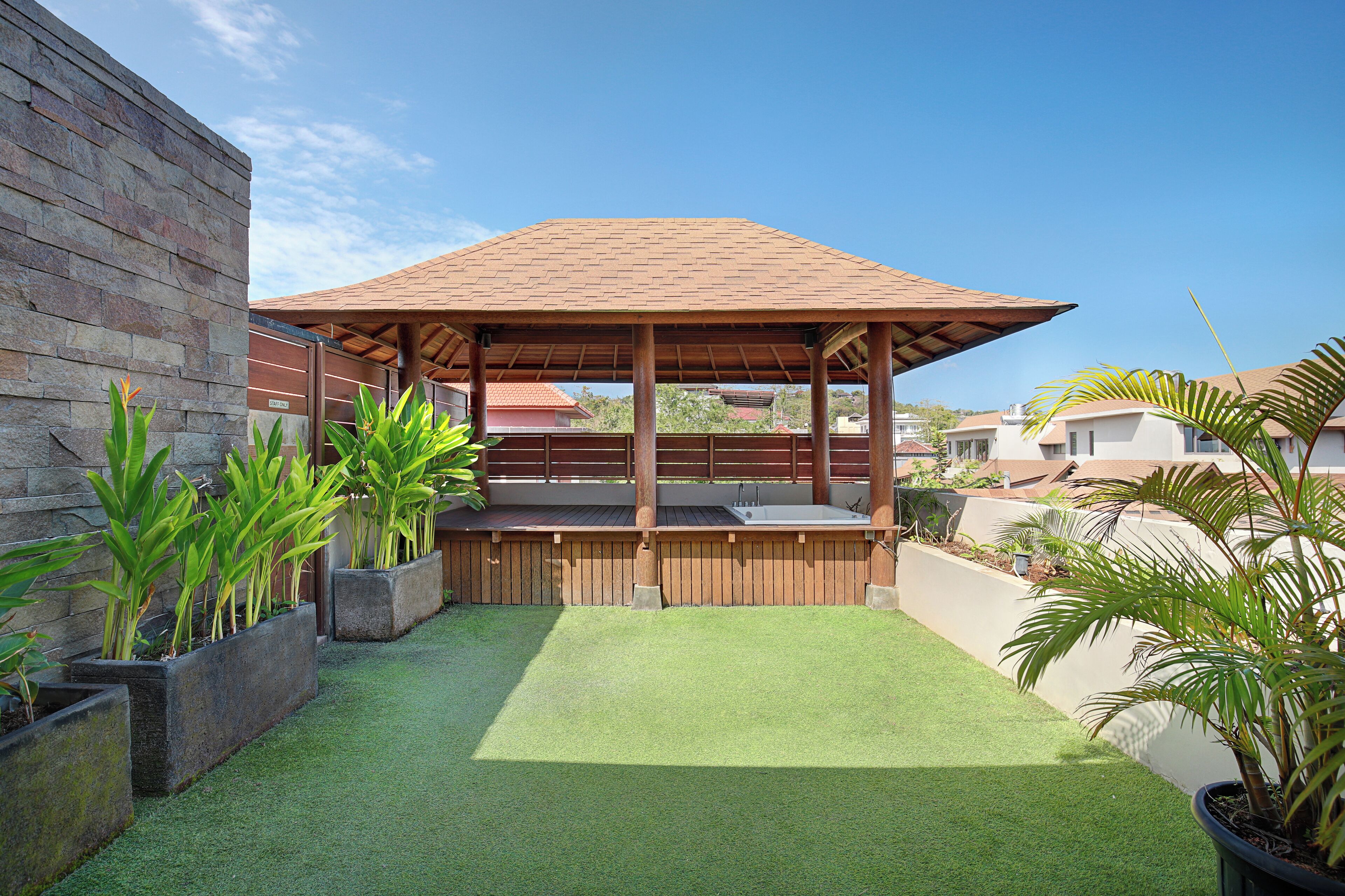 Uma área de varanda coberta com deck de madeira, rodeada por vasos com plantas verdes e um gramado bem cuidado.