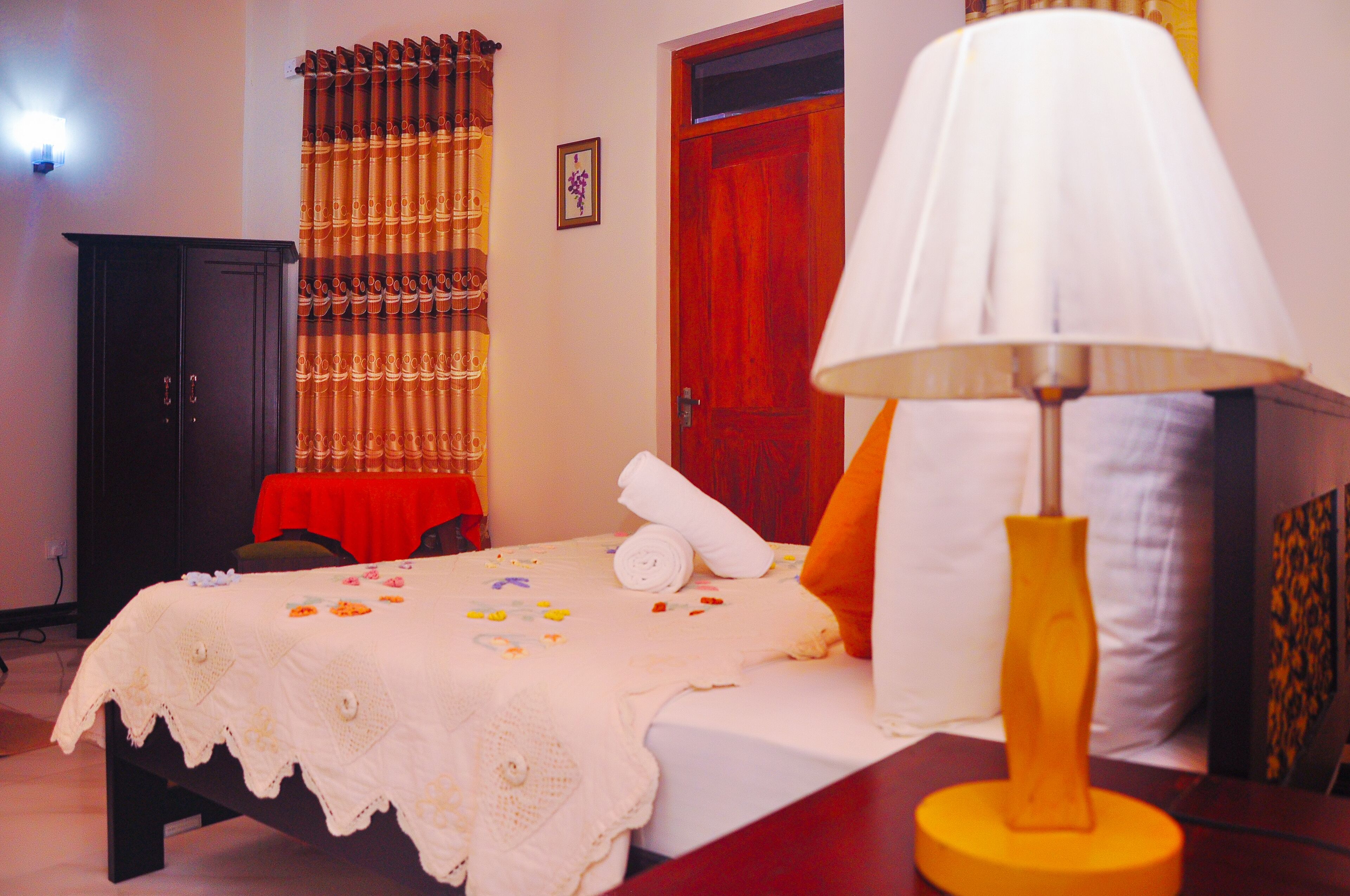 Uma cama bem arrumada com manta e travesseiros brancos, um guarda-roupa de madeira, uma cortina vermelha e um abajur em uma mesa de cabeceira.