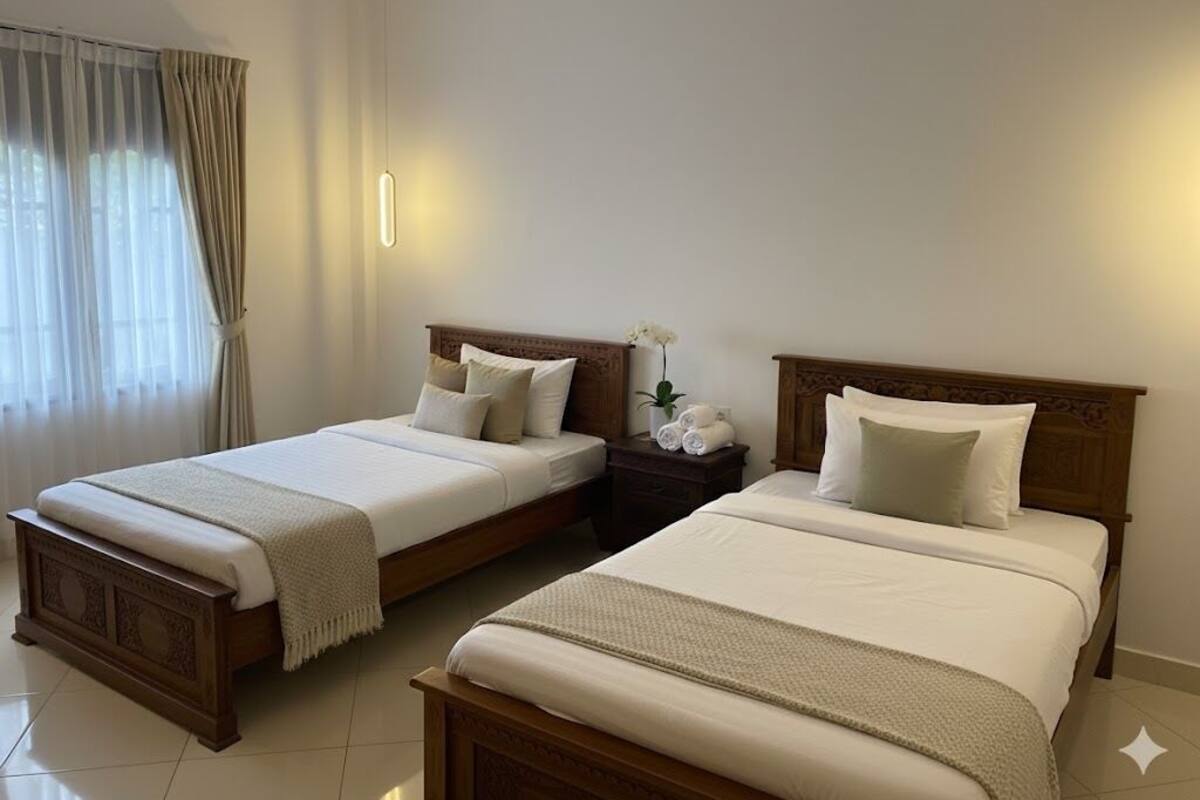 Quarto de hotel com duas camas, uma mesa de cabeceira com um vaso de flores e uma janela com cortinas leves.