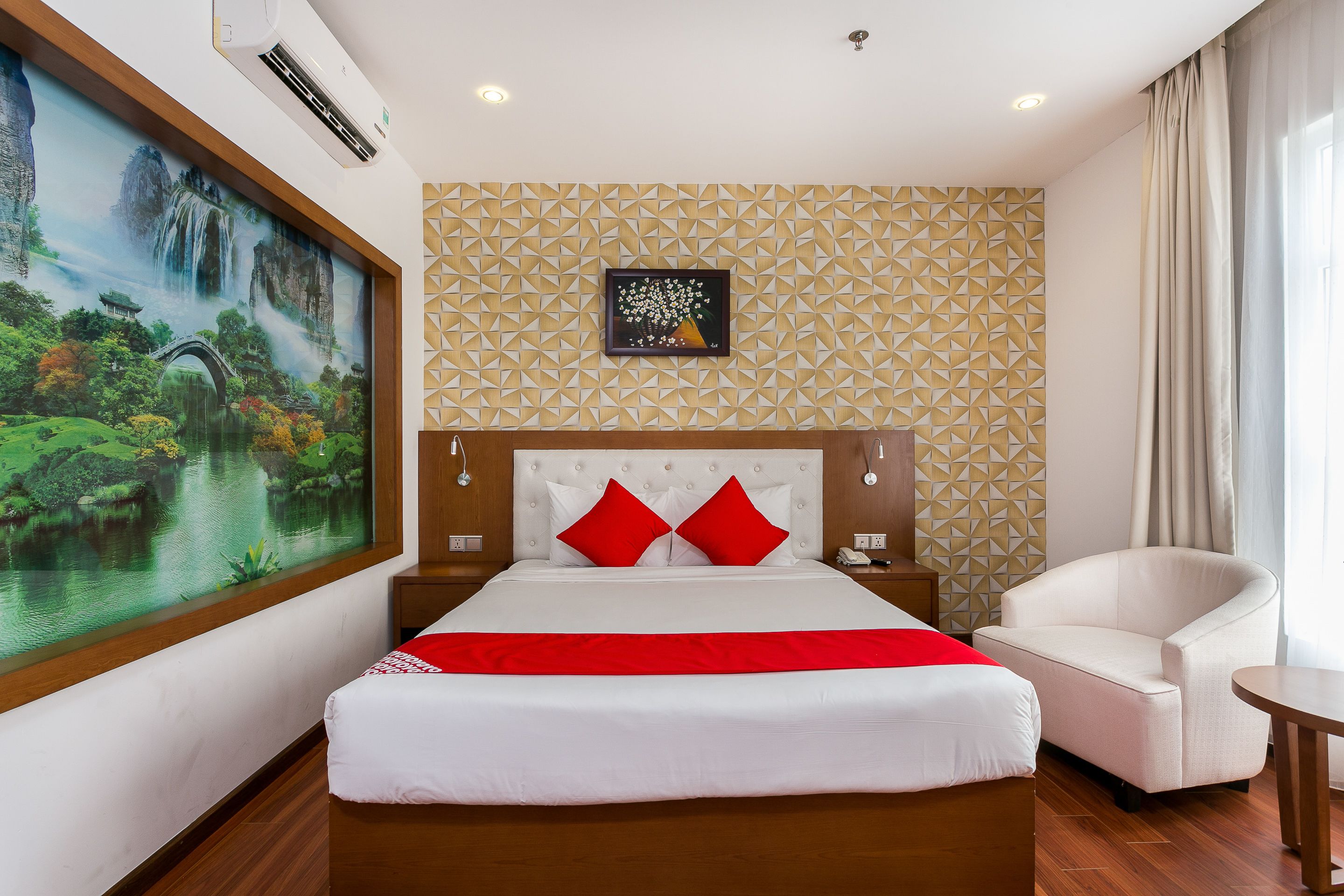 Quarto de hotel com uma cama grande, uma cadeira, uma mesinha, um quadro e parede com padrão geométrico.