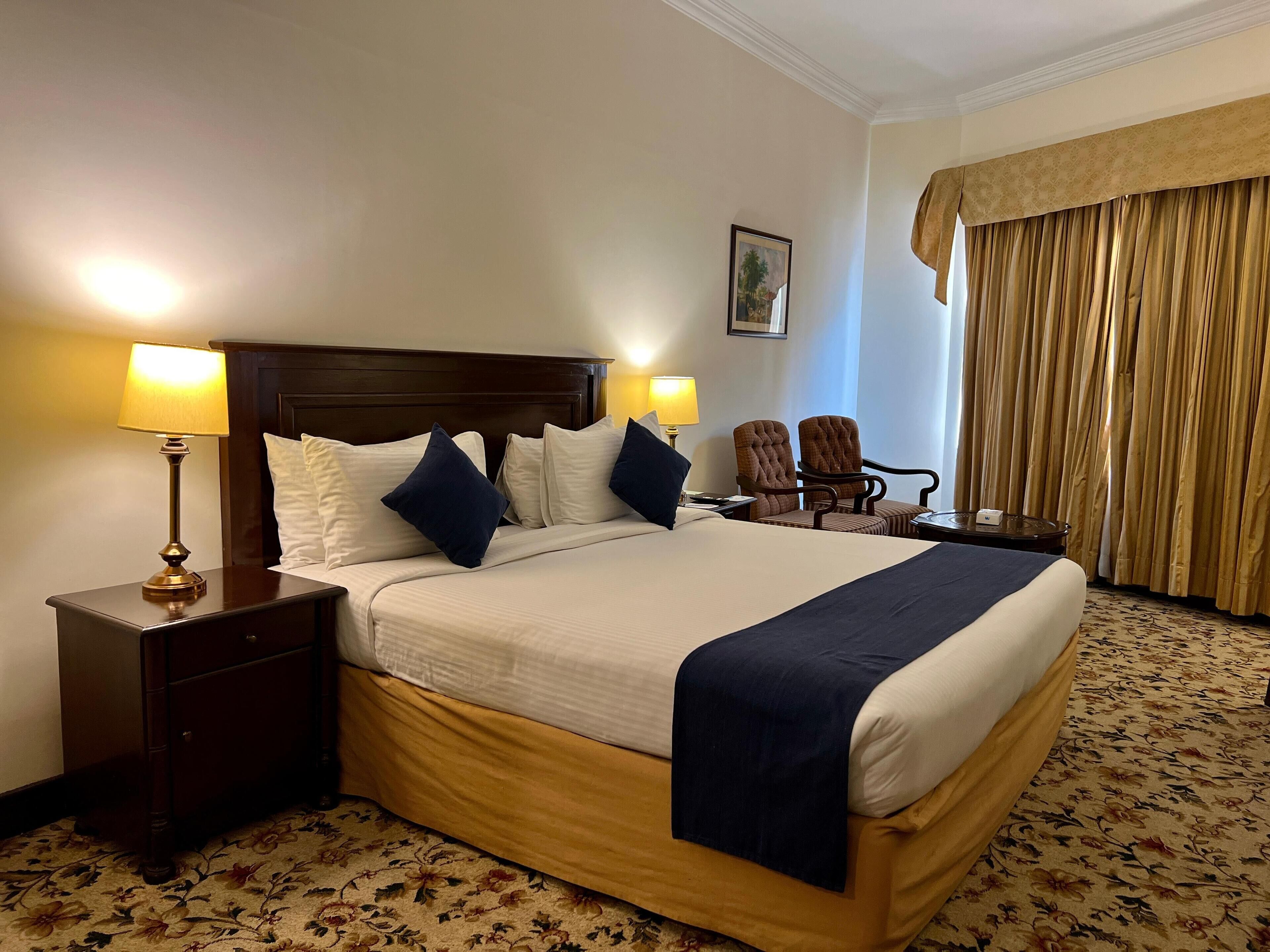 Quarto de hotel com uma cama grande, duas luminárias de cabeceira, uma cadeira, uma escrivaninha e uma janela com cortinas.