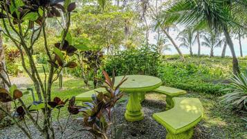 Um jardim com uma mesa e banco de pedra, cercado por plantas e árvores diversas.