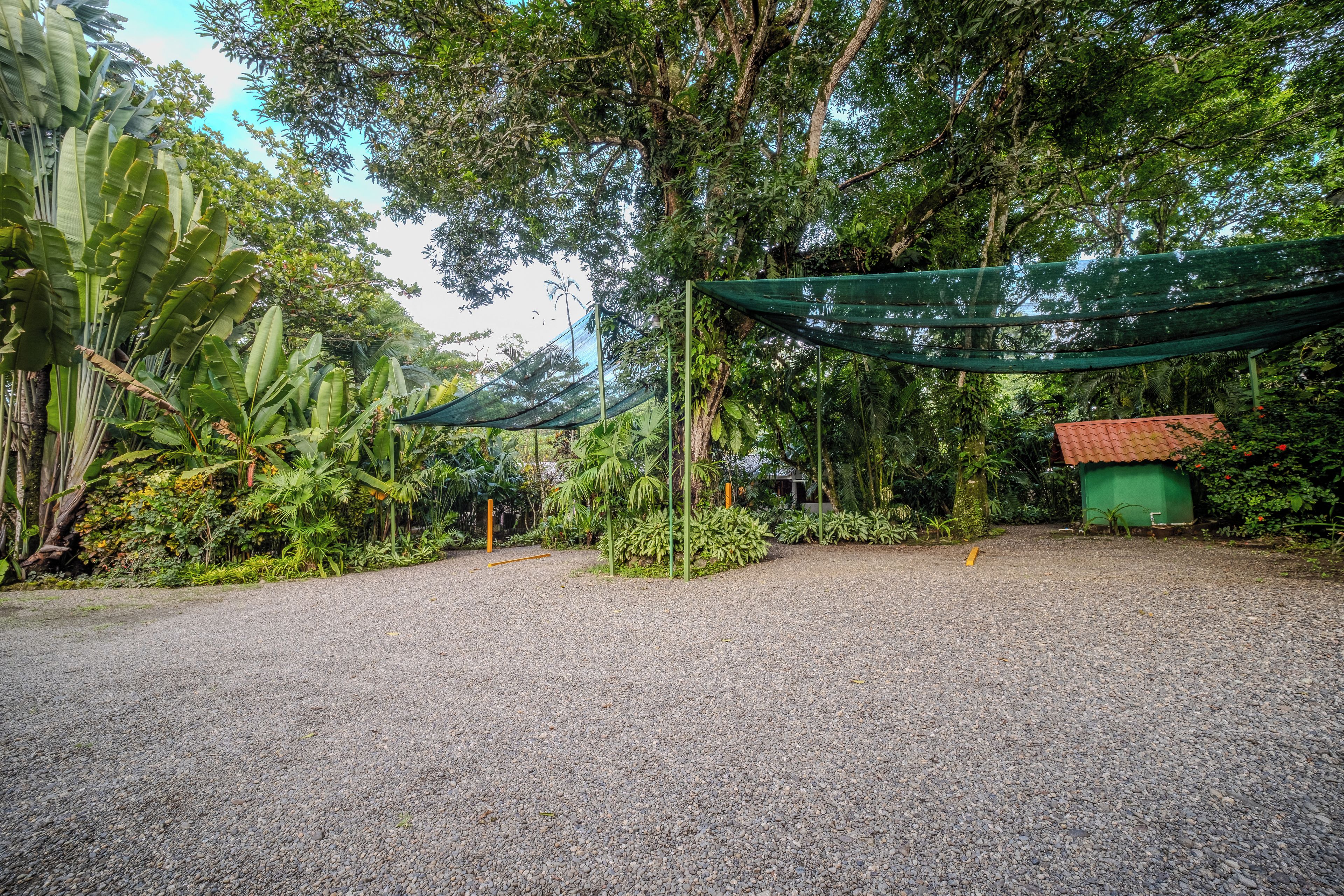 Uma trilha de cascalho leva a um edifício com telhado vermelho, cercado por vegetação tropical densa e sob copas verdes.
