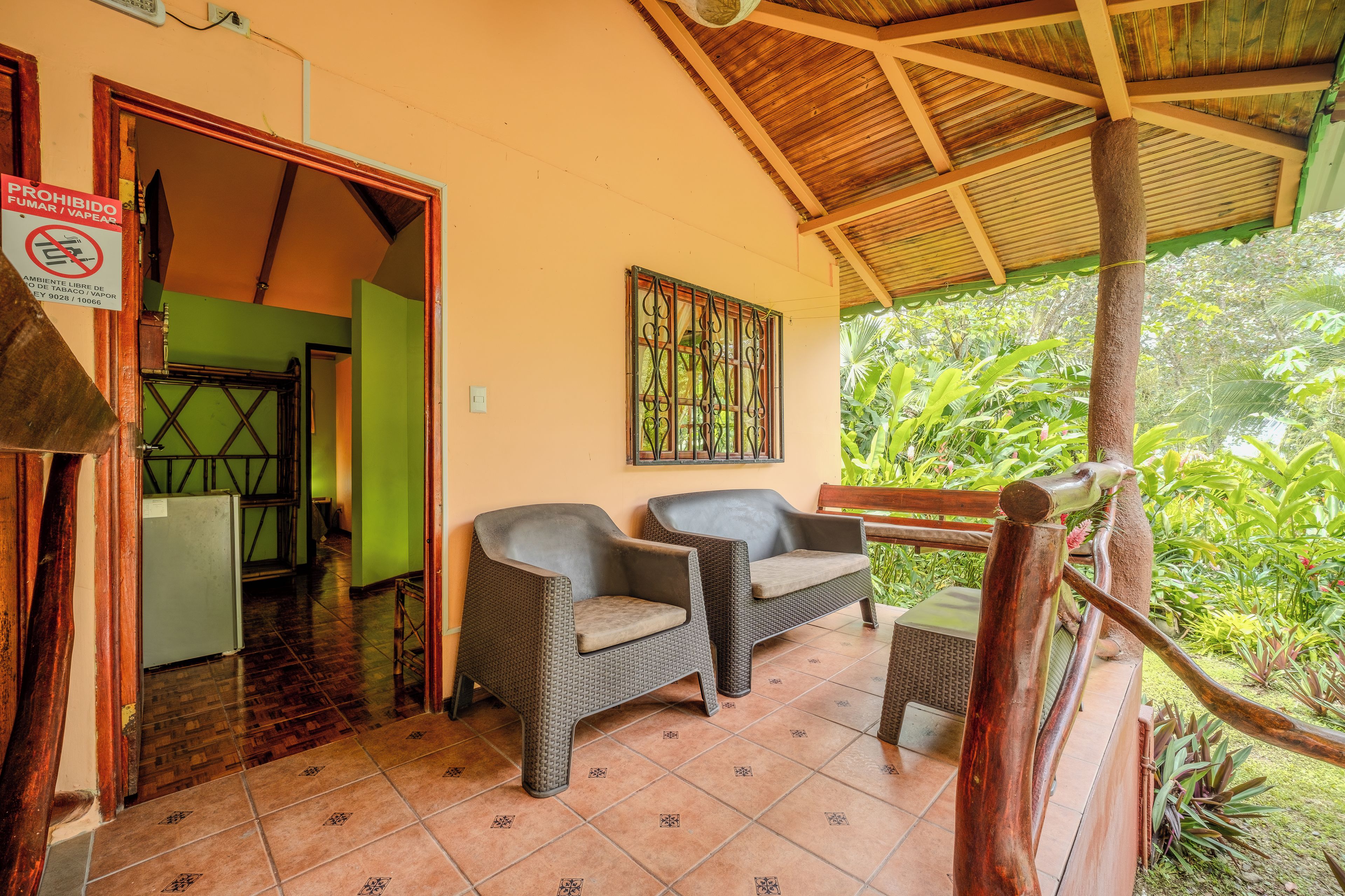 Uma área de estar coberta ao ar livre, com móveis de vime, grade de madeira e vista para vegetação exuberante.