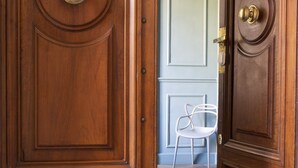 Uma porta de madeira com maçaneta de latão, dando acesso a um quarto com parede azul clara e uma cadeira branca.
