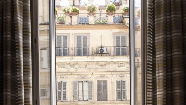 Quarto com cama, um banquinho e vista para um edifício com varandas e janelas.